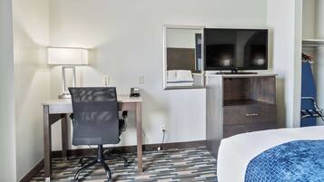 Quarto de hotel com cama, escrivaninha, cadeira, televisão e espelho.