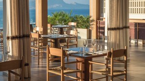 Um restaurante com mesas, cadeiras, janelas amplas e vista para o mar.