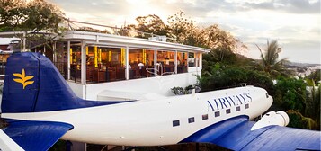 Um avião vintage da Airways com área de passageiros coberta, rodeado por vegetação e vista para a paisagem urbana.
