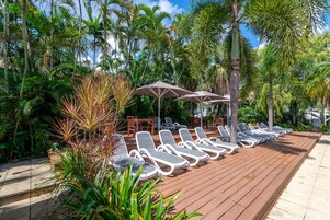 Um deck de madeira com espreguiçadeiras e guarda-sóis, rodeado por vegetação tropical exuberante.