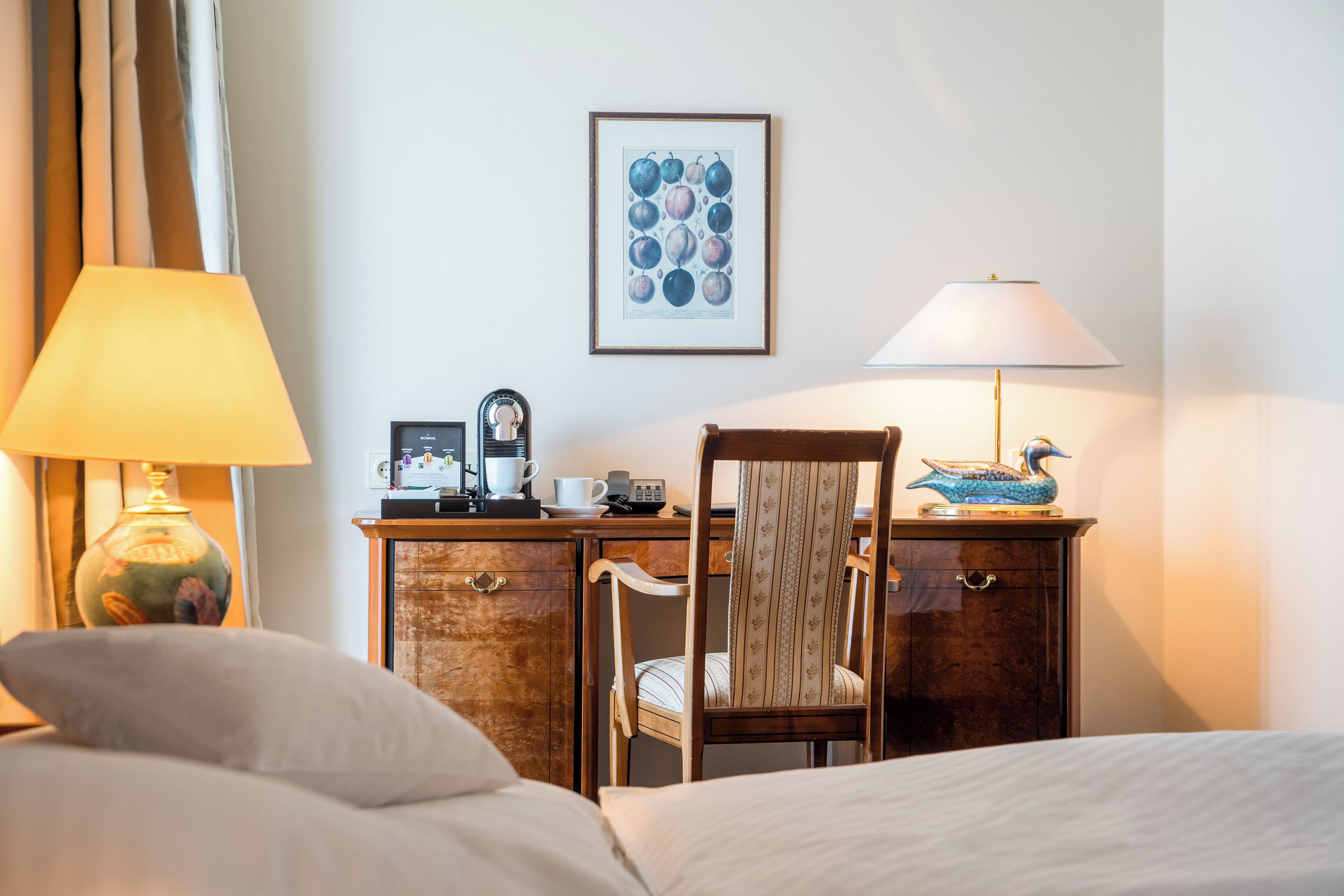 Quarto de hotel com uma cama, uma escrivaninha com abajur, uma cadeira e um quadro na parede.