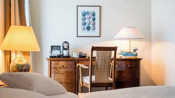 Quarto de hotel com uma cama, uma escrivaninha com abajur, uma cadeira e um quadro na parede.