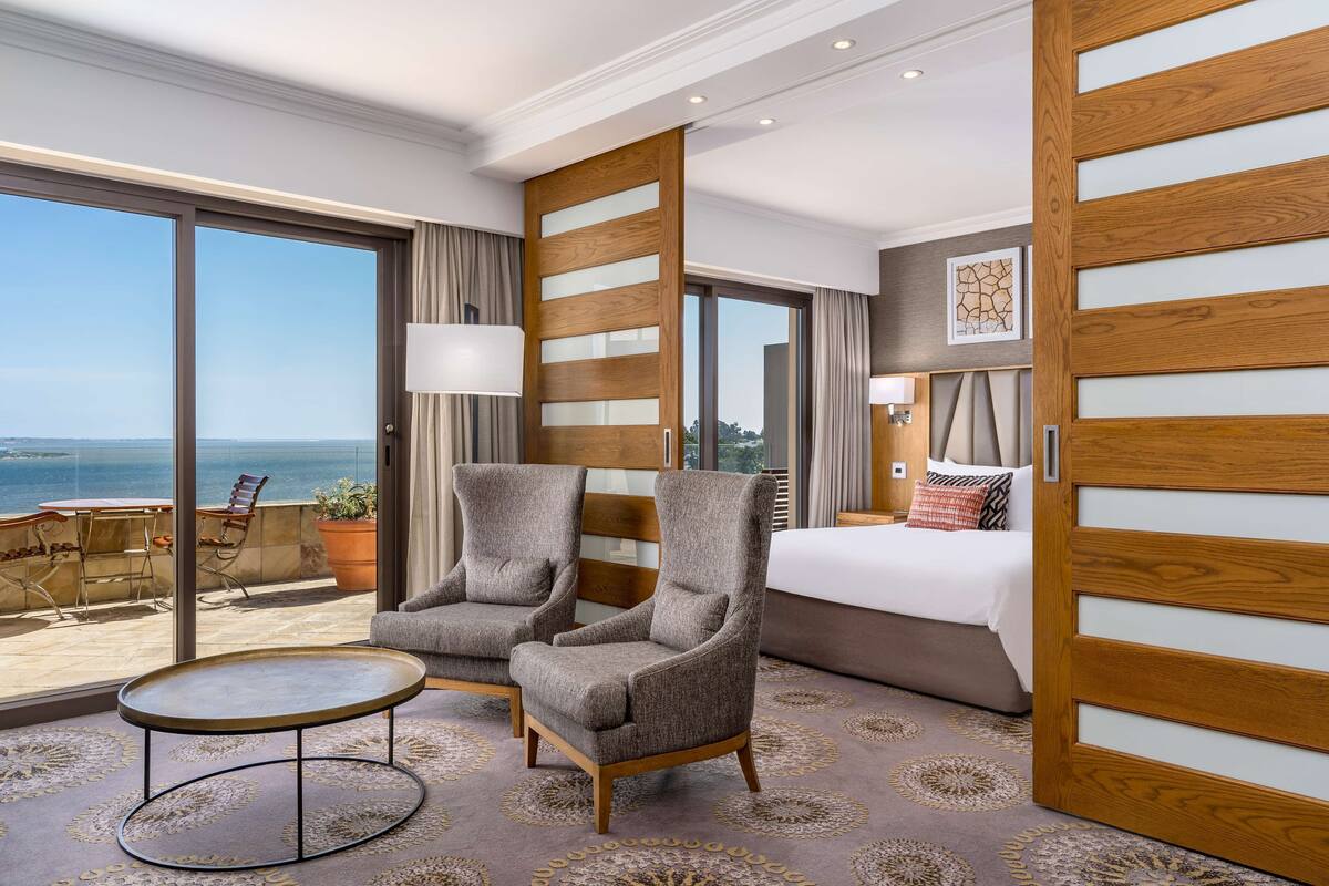Quarto de hotel com uma janela grande, uma cama, duas poltronas, uma mesa pequena e um vaso de planta.