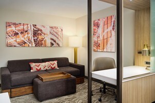 Quarto de hotel moderno com sofá cinzento, mesa lateral de madeira, escrivaninha e cadeira, e arte abstrata nas paredes.