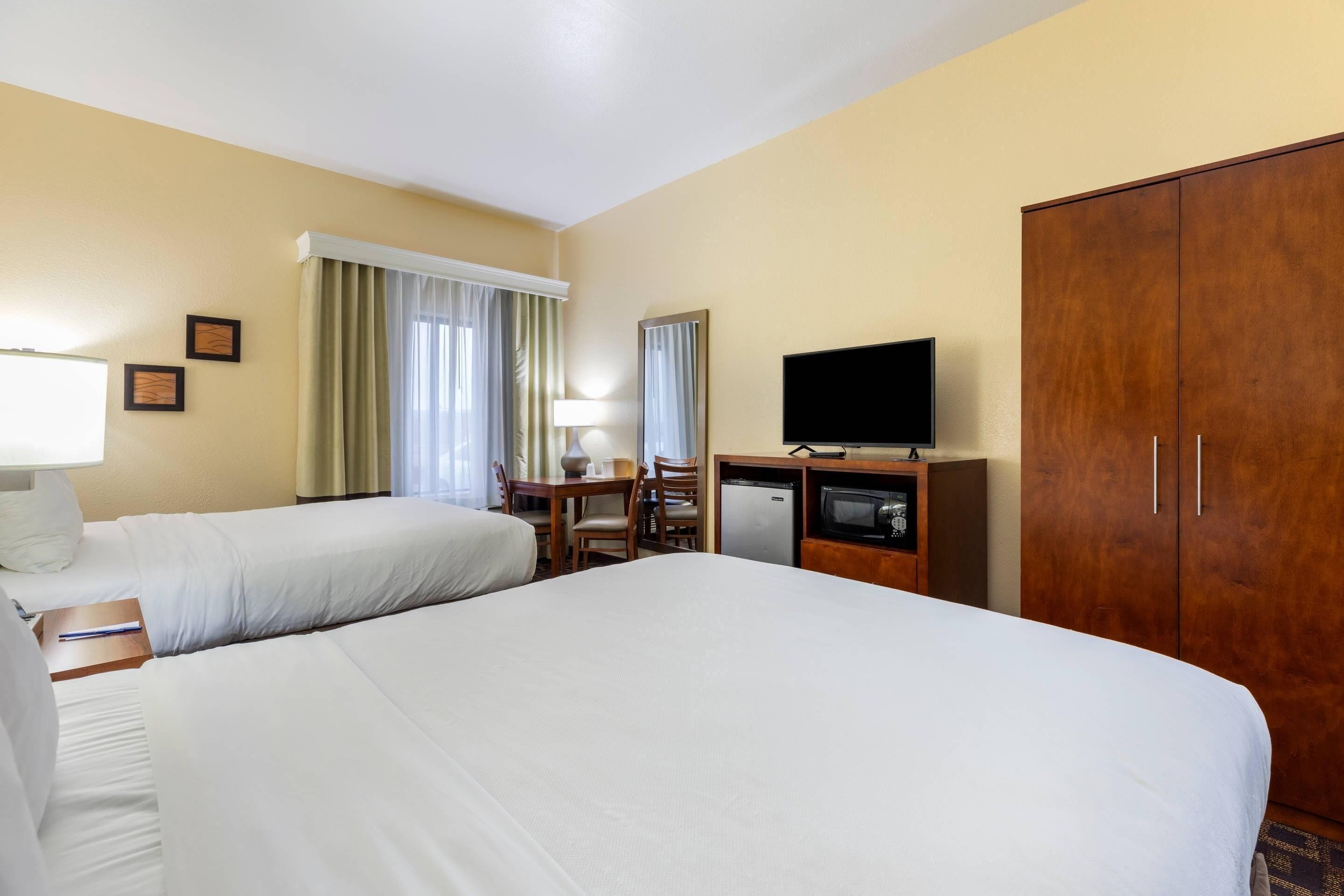 Quarto de hotel com duas camas, uma escrivaninha com cadeira, televisão, guarda-roupa e janela com cortinas.