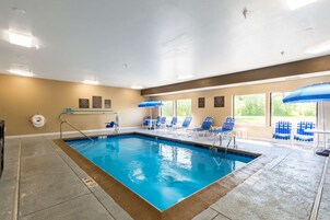 Piscina interior com cadeiras e guarda-sóis azuis, cadeira de salva-vidas e ampla vista para o exterior através de grandes janelas.