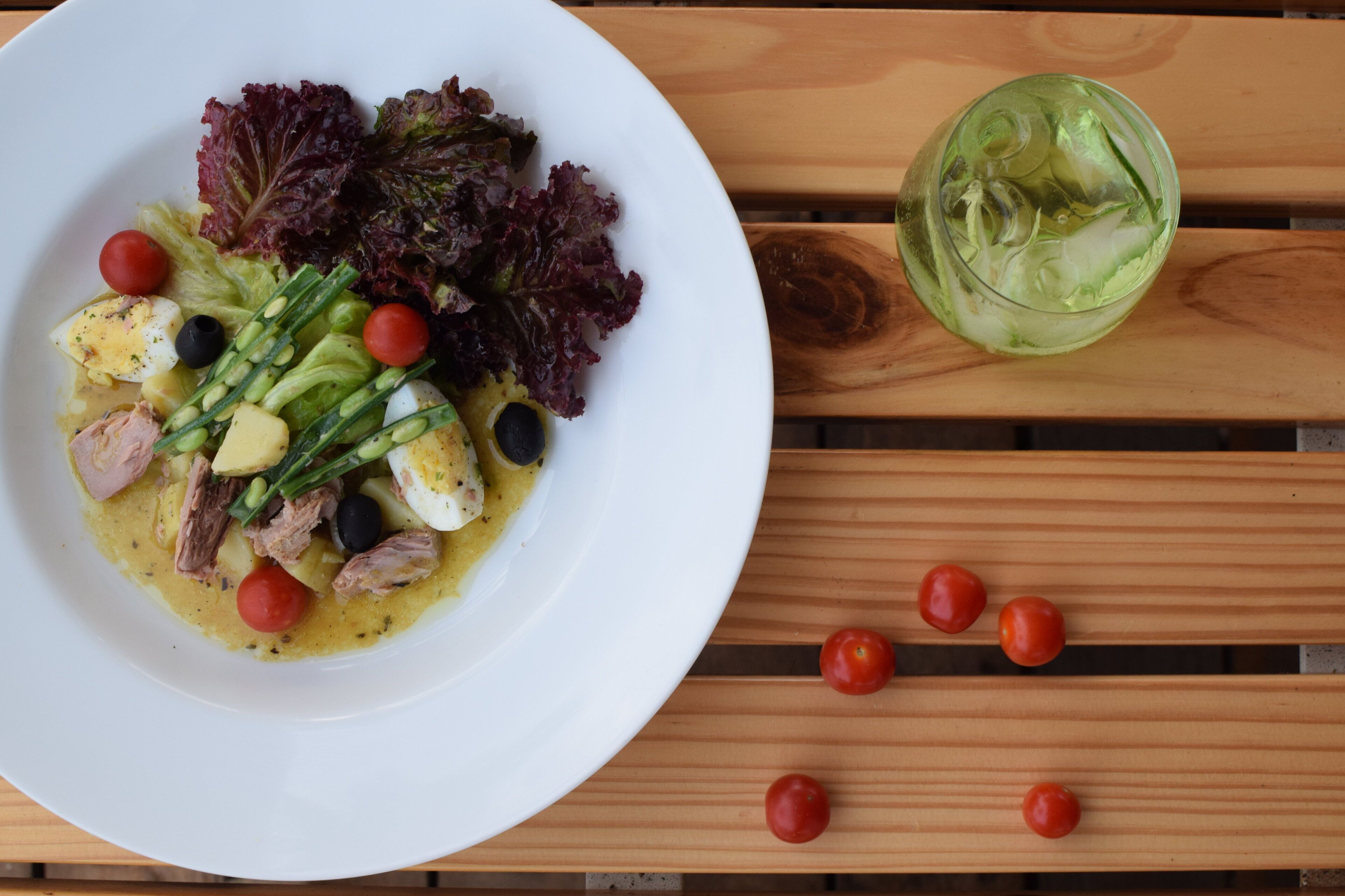Um prato branco com salada, um copo de bebida verde e tomates cereja espalhados sobre uma mesa de madeira.