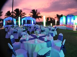 Espaço para eventos ao ar livre com mesas redondas dispostas para refeições, decorado com toalhas brancas e fitas azuis, e iluminado por iluminação ambiente.