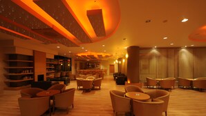 Ampla área de convívio com diversos arranjos de assentos, um bar ao fundo e teto bem iluminado com iluminação decorativa.