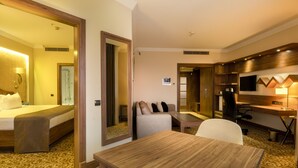 Quarto de hotel com cama, sofá, escrivaninha, cadeira, televisão e espelho.