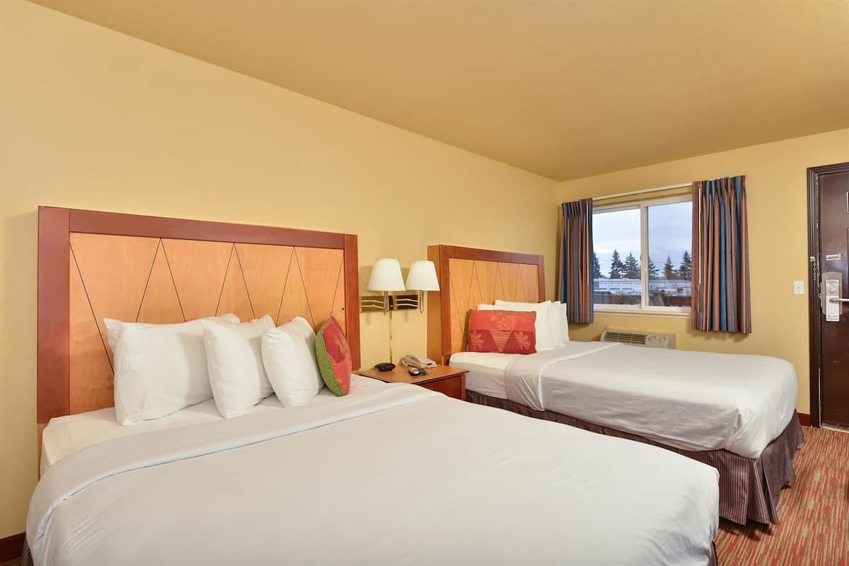 Quarto de hotel com duas camas, uma mesinha de cabeceira, um abajur e uma janela com cortinas.