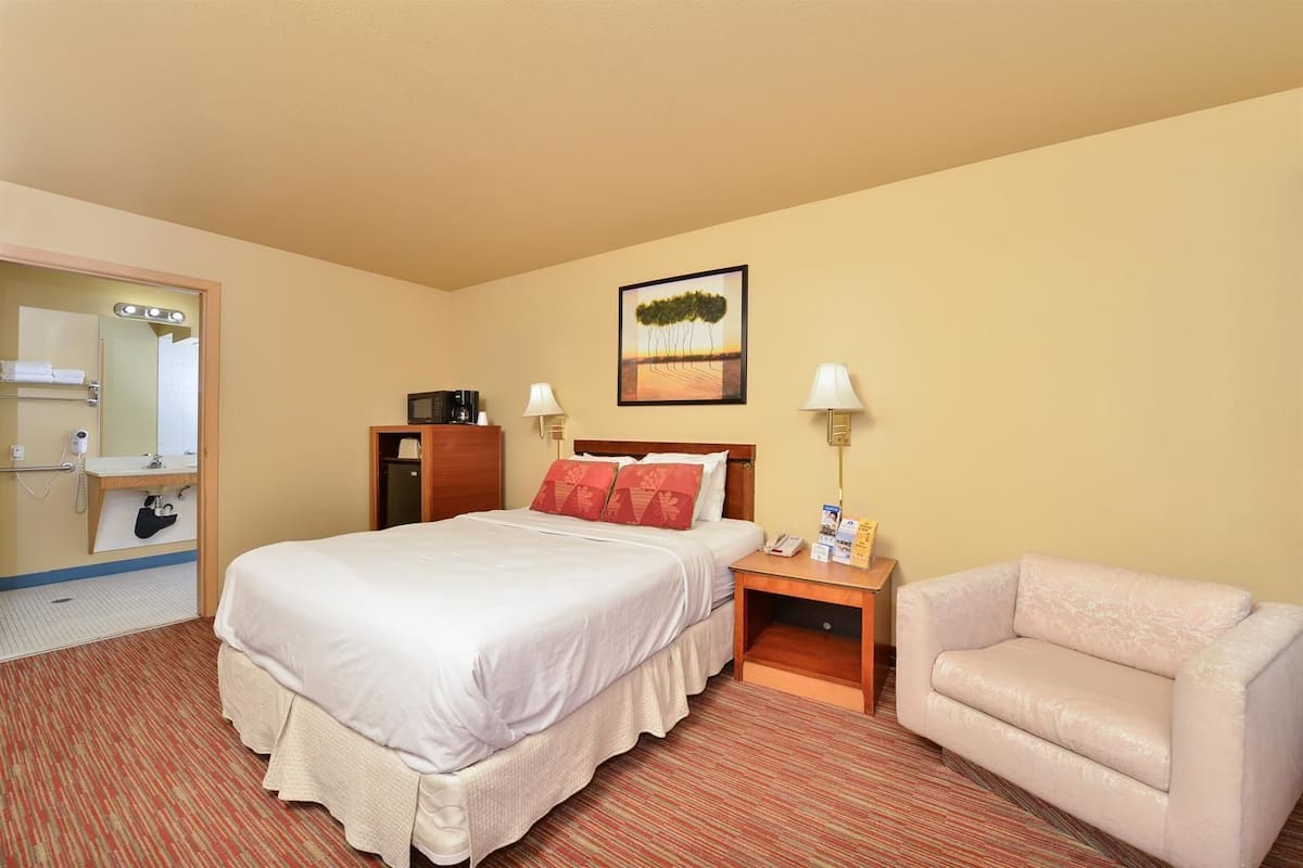 Quarto de hotel com uma cama grande, uma cadeira, uma mesa de cabeceira com abajur, uma mesinha com telefone e folhetos, e um quadro na parede.