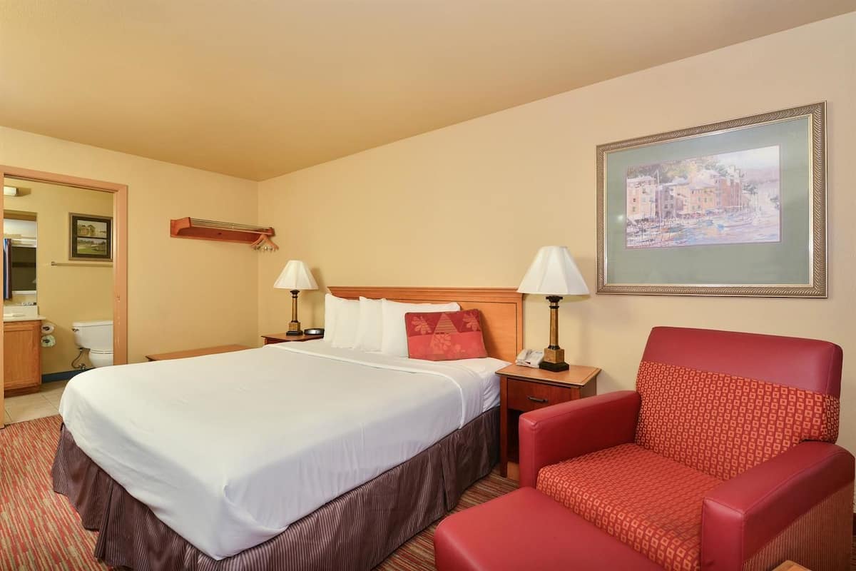 Quarto de hotel com uma cama grande, uma poltrona vermelha, mesa de cabeceira com abajur e um quadro pendurado na parede.