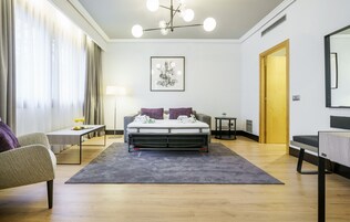 Quarto de hotel moderno com cama, escrivaninha, poltrona, sofá e uma janela grande com cortinas.
