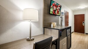 Quarto de hotel com escrivaninha, luminária, televisão fixada na parede, frigobar e uma porta.
