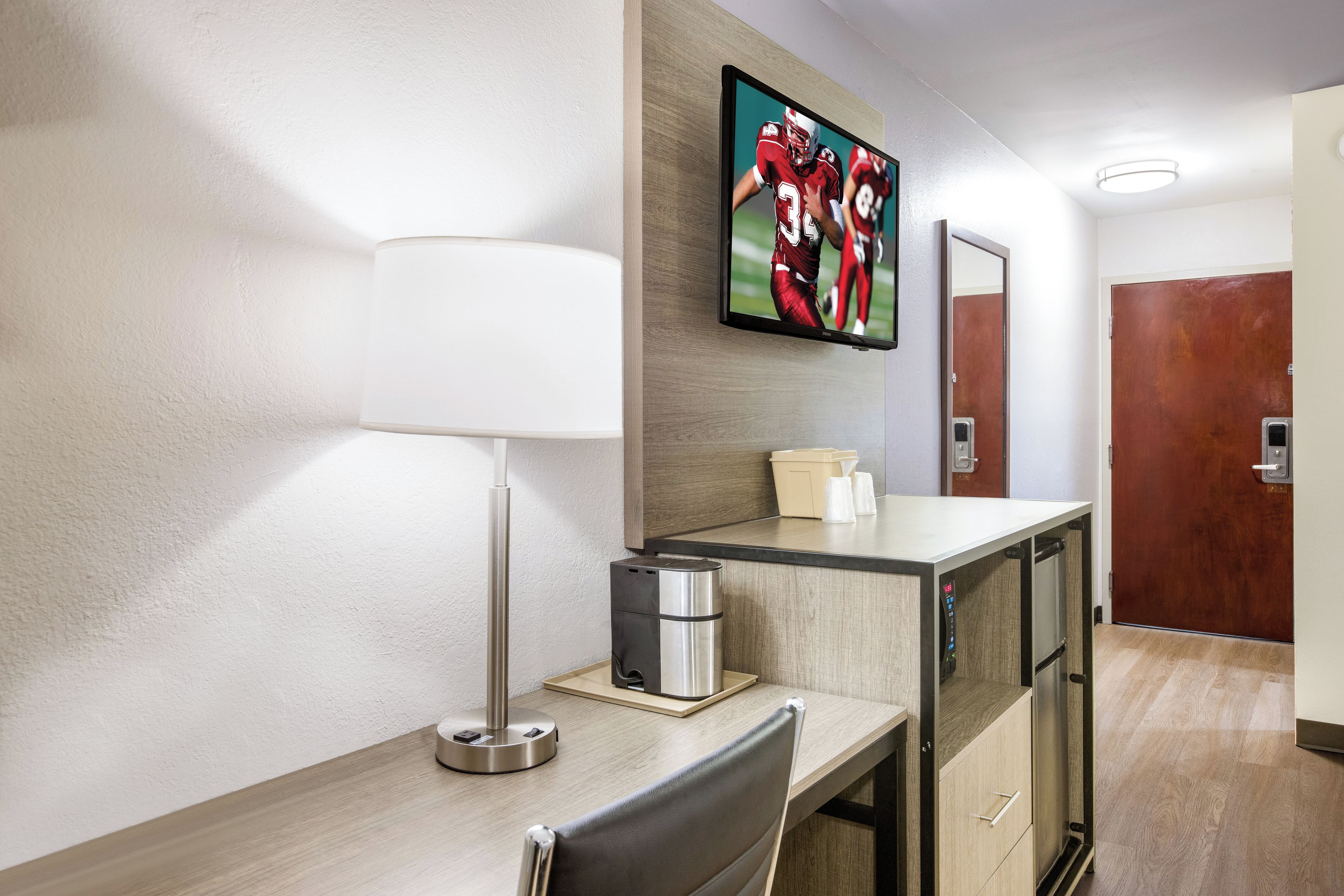 Quarto de hotel com escrivaninha, luminária, televisão na parede e uma porta ao fundo.