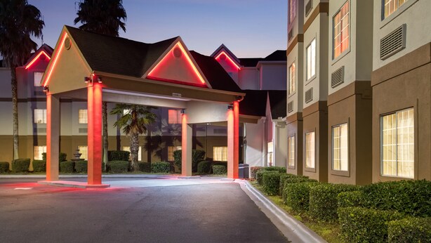 Entrada de hotel com galeria coberta, palmeiras e gramado bem cuidado.