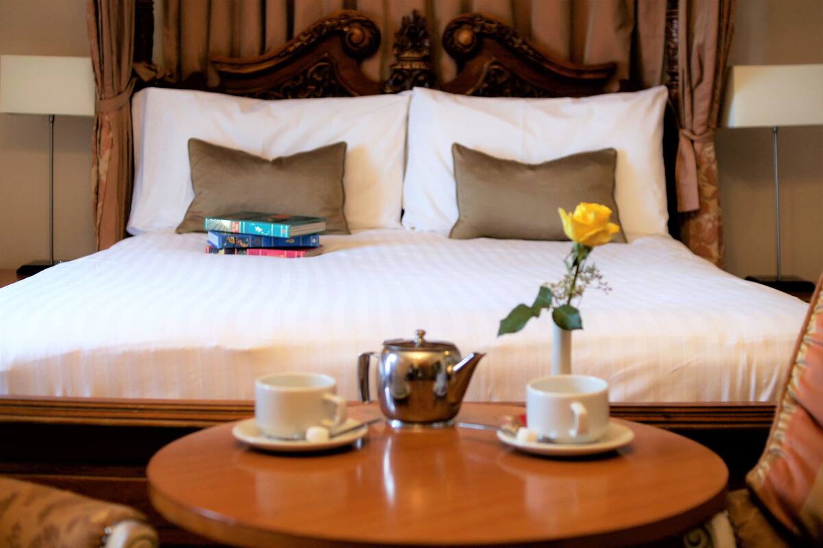 Cama bem arrumada com almofadas, mesa de cabeceira com bule e xícaras, e uma única rosa amarela.