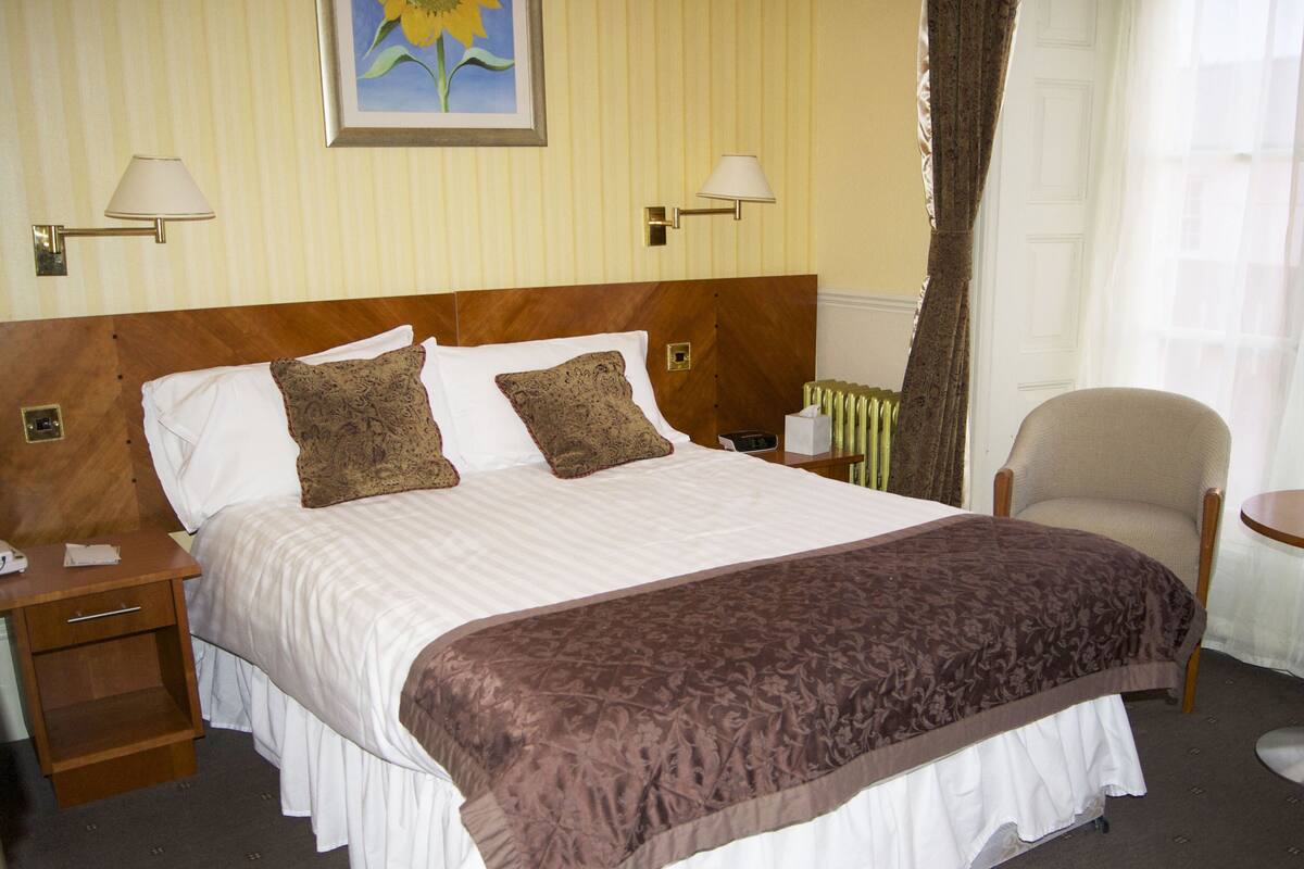 Quarto de hotel com uma cama grande, duas luminárias de cabeceira, uma poltrona, uma mesinha e um radiador.