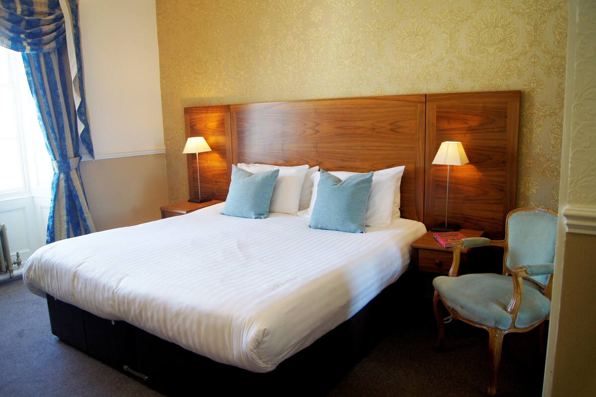 Quarto de hotel com uma cama grande, duas luminárias de cabeceira, uma cadeira e uma mesinha de cabeceira.