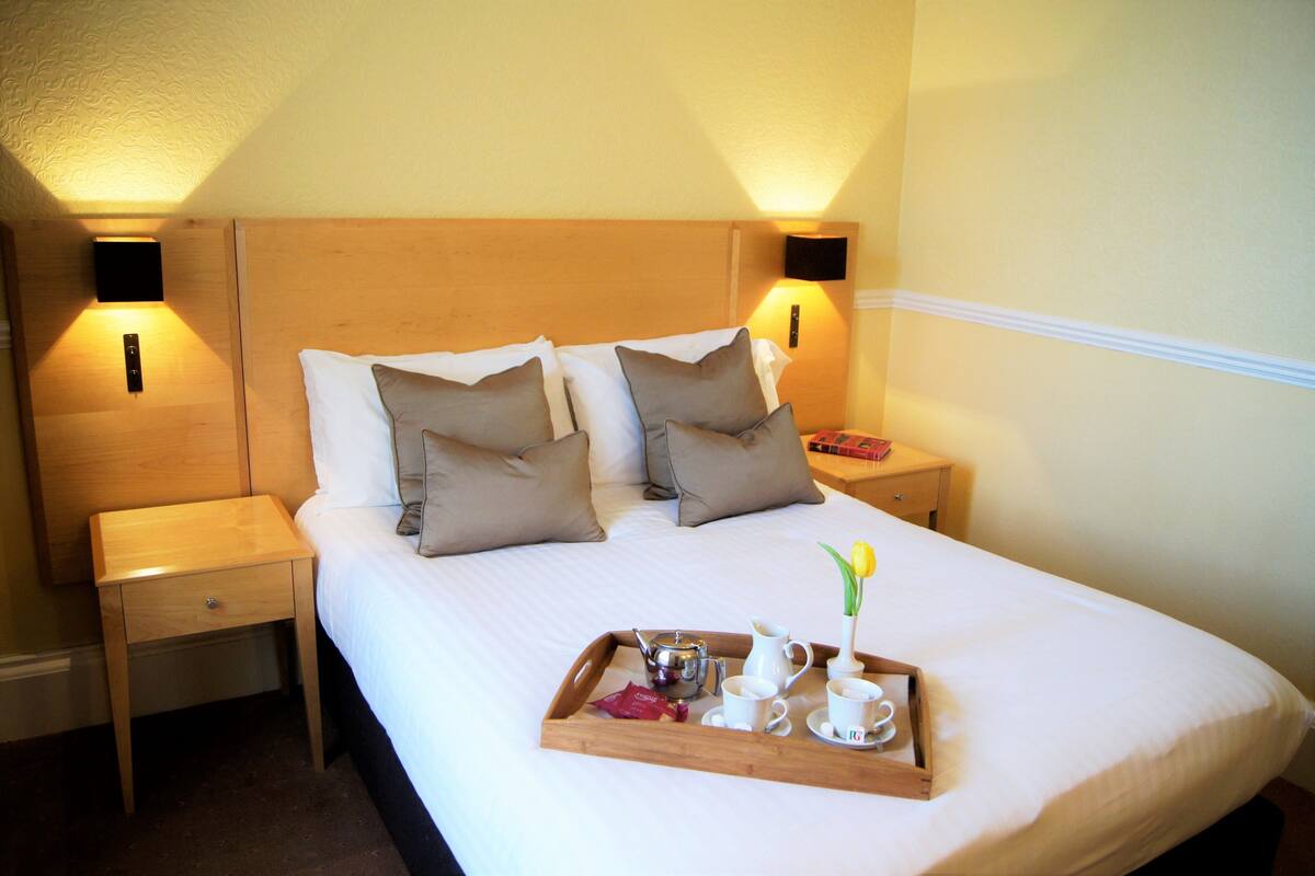 Cama bem arrumada com travesseiros, mesa de cabeceira e bandeja com chá e uma flor.