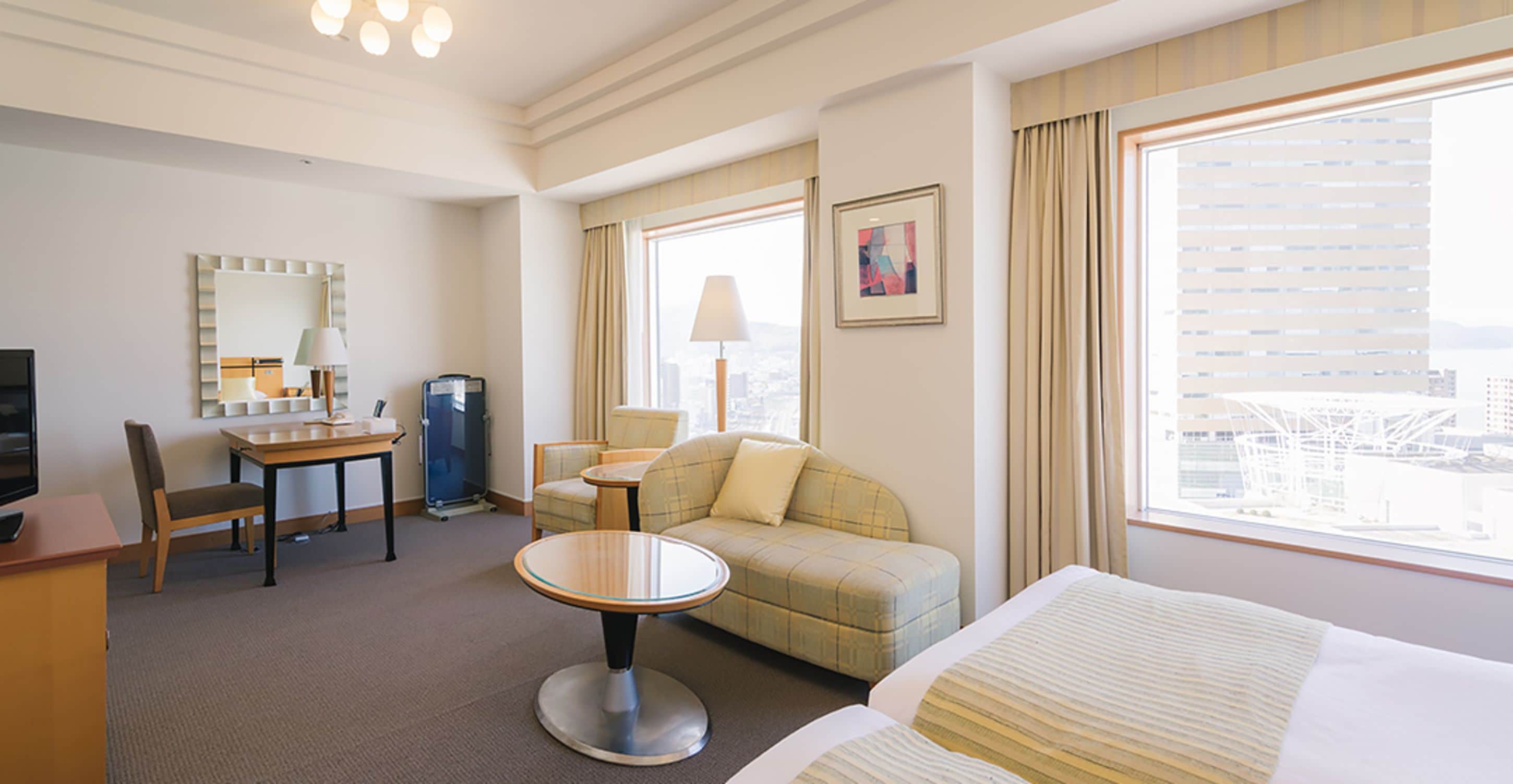 Quarto de hotel com cama, sofá, escrivaninha com cadeira, mesa pequena, televisão e janela com vista para a cidade.