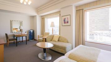 Quarto de hotel com cama, sofá, escrivaninha com cadeira, mesa pequena, televisão e janela com vista para a cidade.