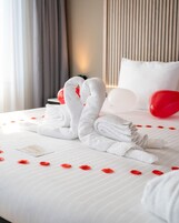 Cama bem arrumada com toalhas brancas dobradas, pétalas de rosa vermelha e um cartão de boas-vindas.