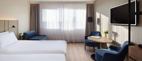 Quarto de hotel com duas camas, uma escrivaninha com cadeira, TV fixada na parede e uma janela grande com cortinas.