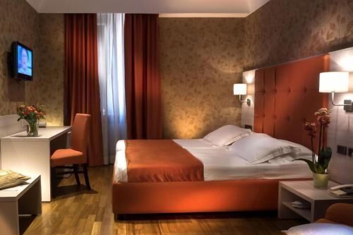 Quarto de hotel com cama, escrivaninha, cadeira e televisão.