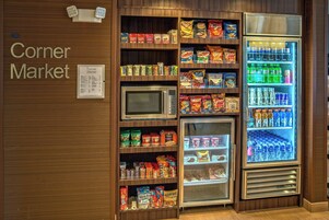 Uma mercearia de esquina com prateleiras cheias de lanches, um micro-ondas e um refrigerador para bebidas.