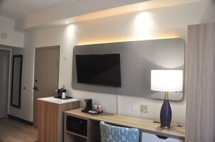 Quarto de hotel com TV de tela plana na parede, escrivaninha com luminária, cafeteira e uma cadeira.