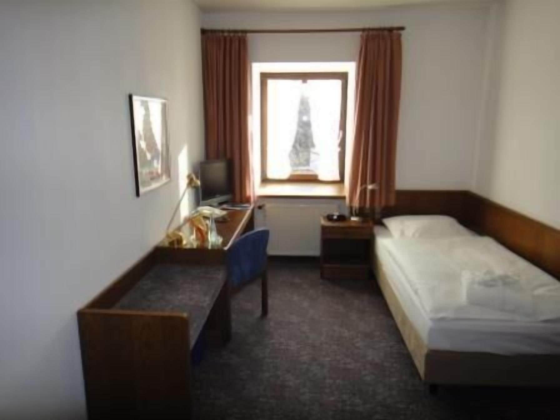 Quarto de hotel com uma cama de solteiro, uma escrivaninha com cadeira, uma mesinha, um criado-mudo e uma janela com cortinas.