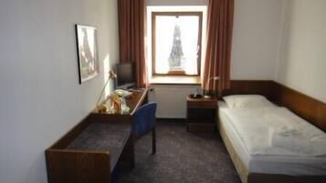 Quarto de hotel com uma cama de solteiro, uma escrivaninha com cadeira, uma mesinha, um criado-mudo e uma janela com cortinas.