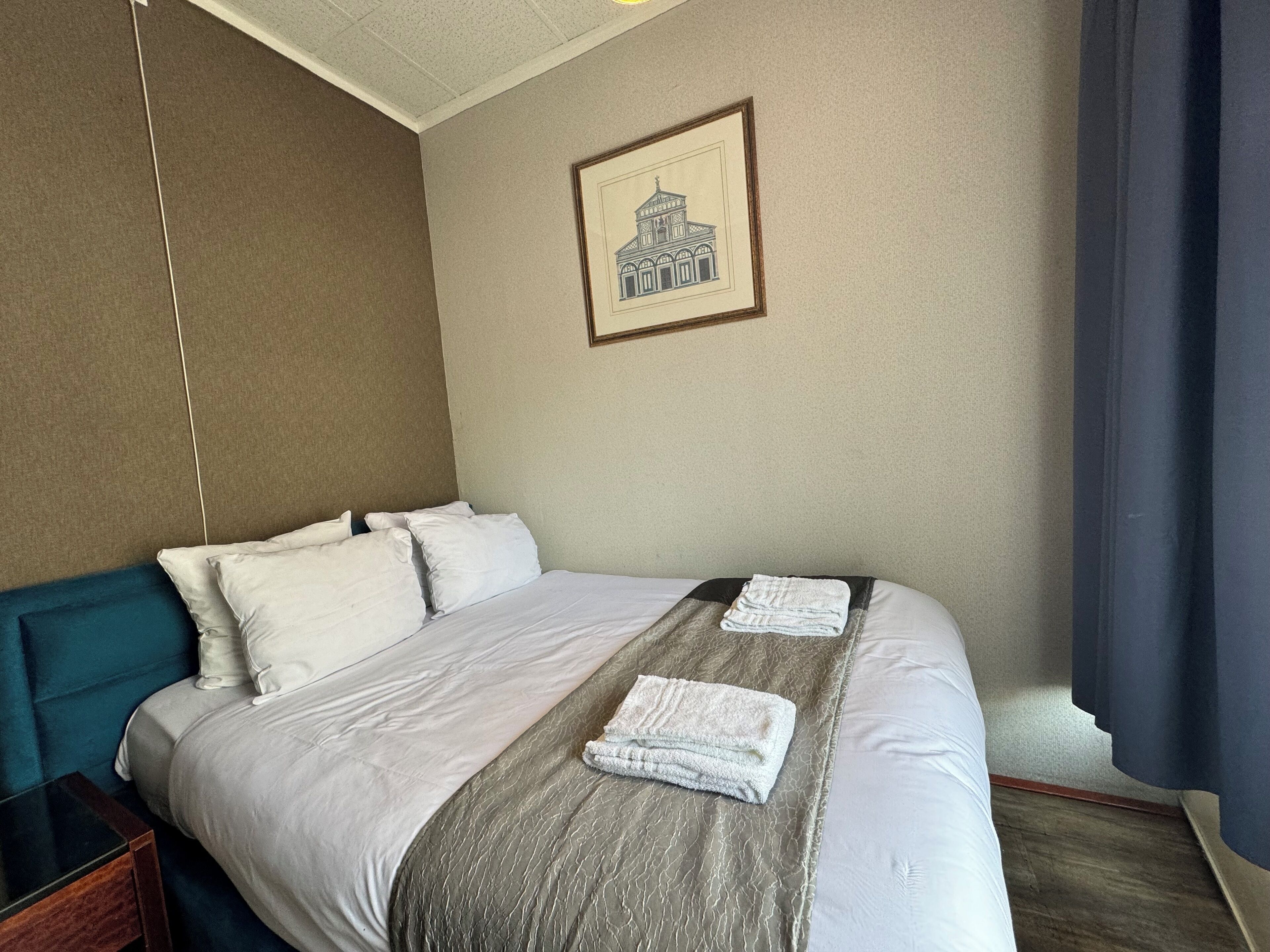 Quarto de hotel com uma cama grande, roupa de cama branca, um edredom cinzento e um quadro na parede.