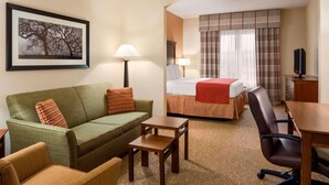 Quarto de hotel com sofá verde, uma mesinha, poltrona marrom, cama com manta vermelha e televisão sobre um suporte de madeira.