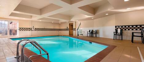 Piscina interna com borda de azulejos quadriculada, deck de madeira e teto com padrões geométricos.