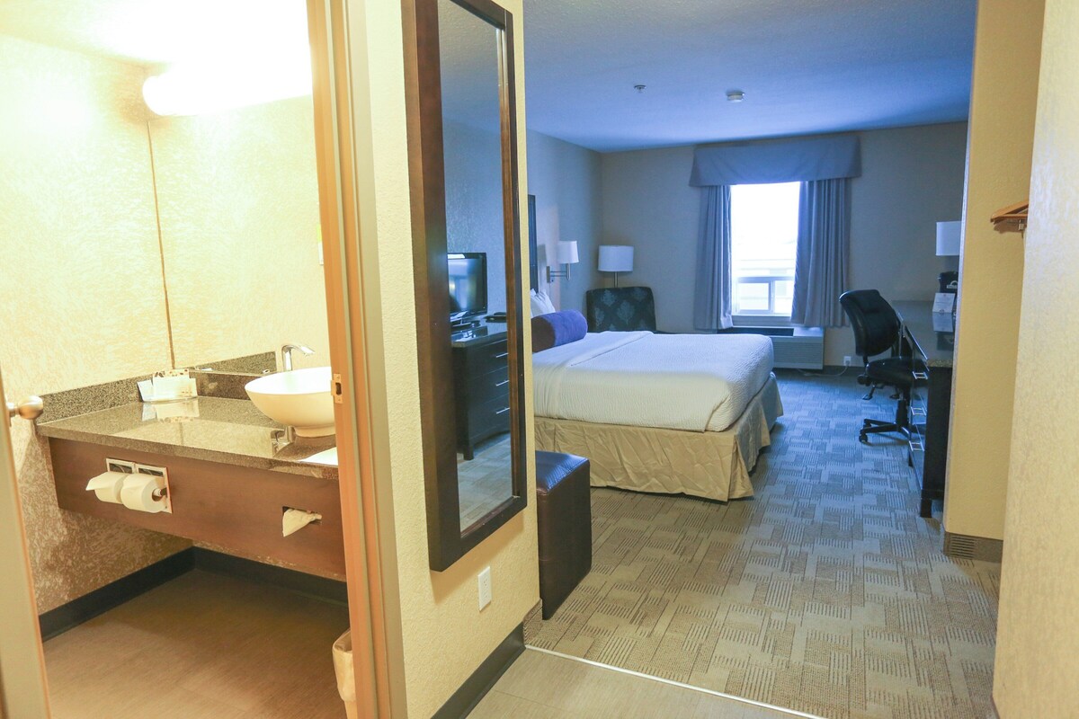 Quarto de hotel com cama, escrivaninha, cadeira, televisão, banheiro com pia e espelho, e janela com cortinas.