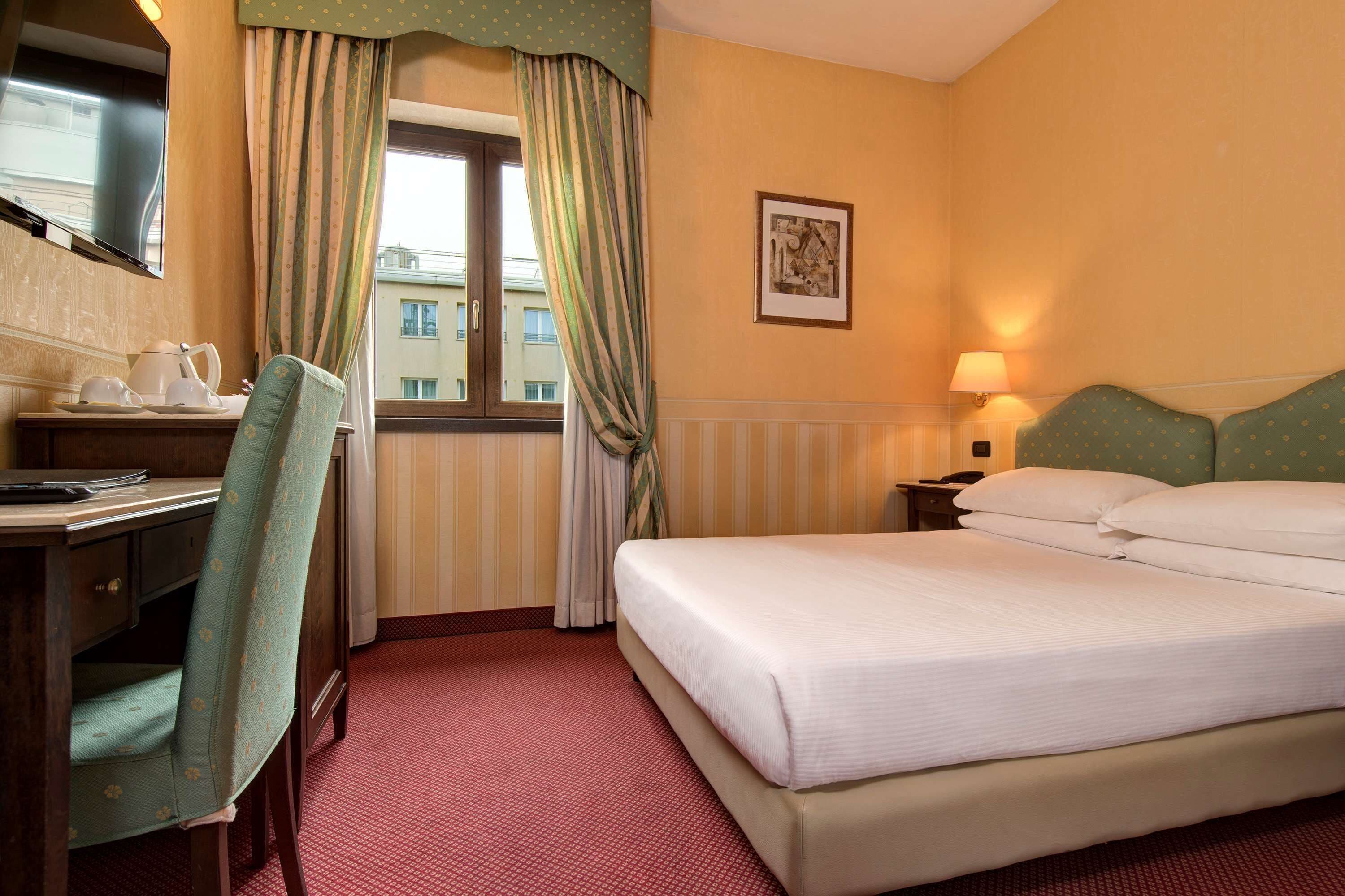 Quarto de hotel com cama, escrivaninha, cadeira e janela com cortinas.