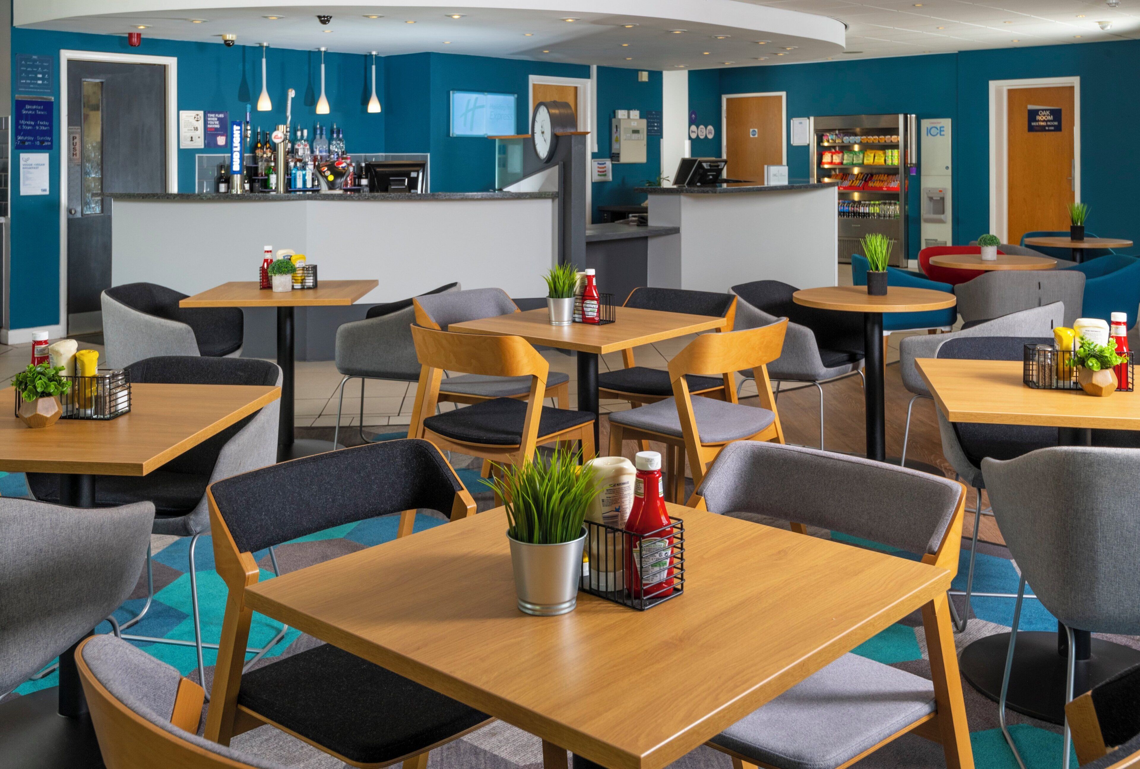 Uma sala de jantar de hotel com várias mesas e cadeiras, um bar e um relógio na parede.