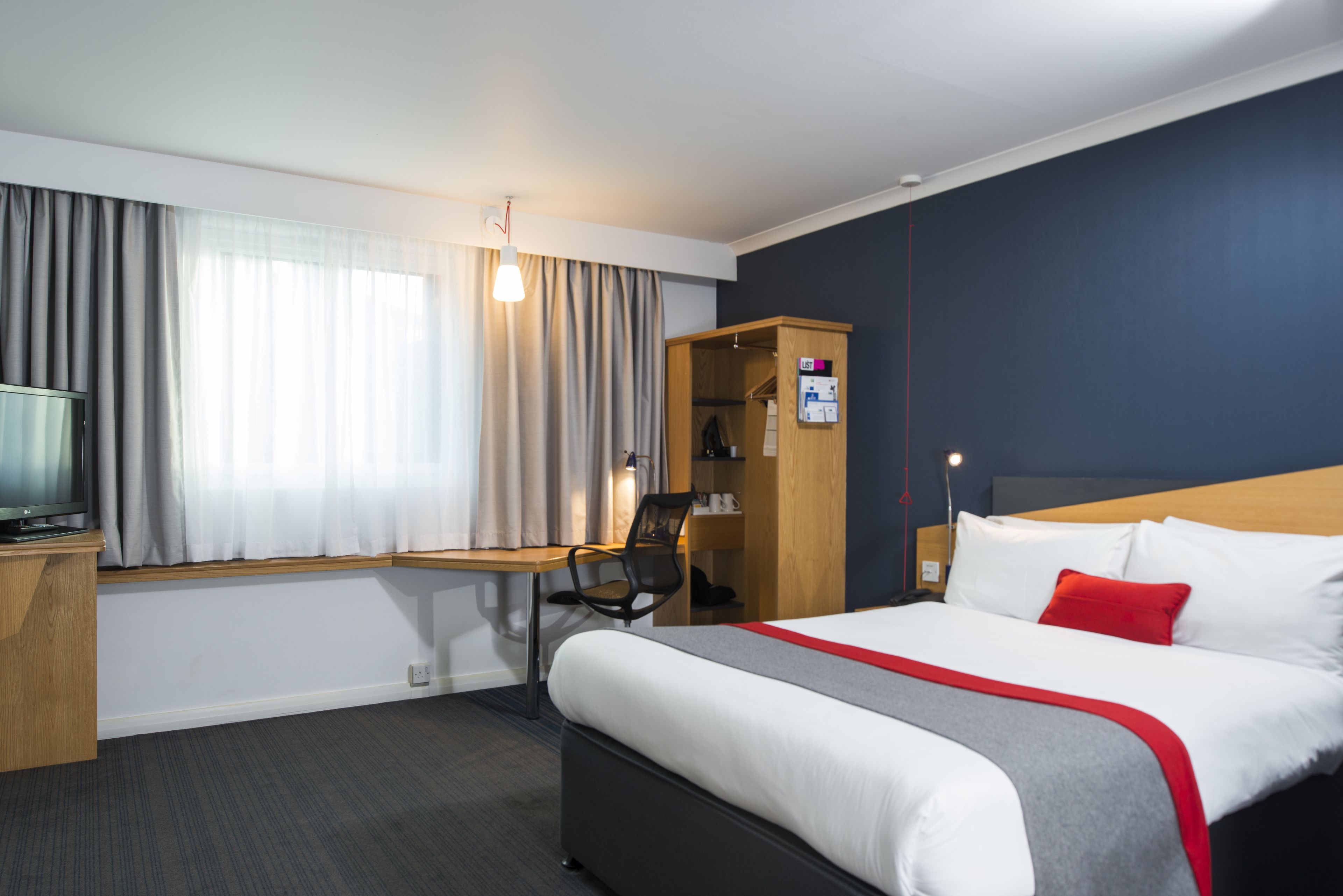 Quarto de hotel com cama, escrivaninha, cadeira, televisão e armário.