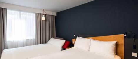 Quarto de hotel com duas camas, cabeceira de madeira, mesa de cabeceira com abajur e janela com cortinas.
