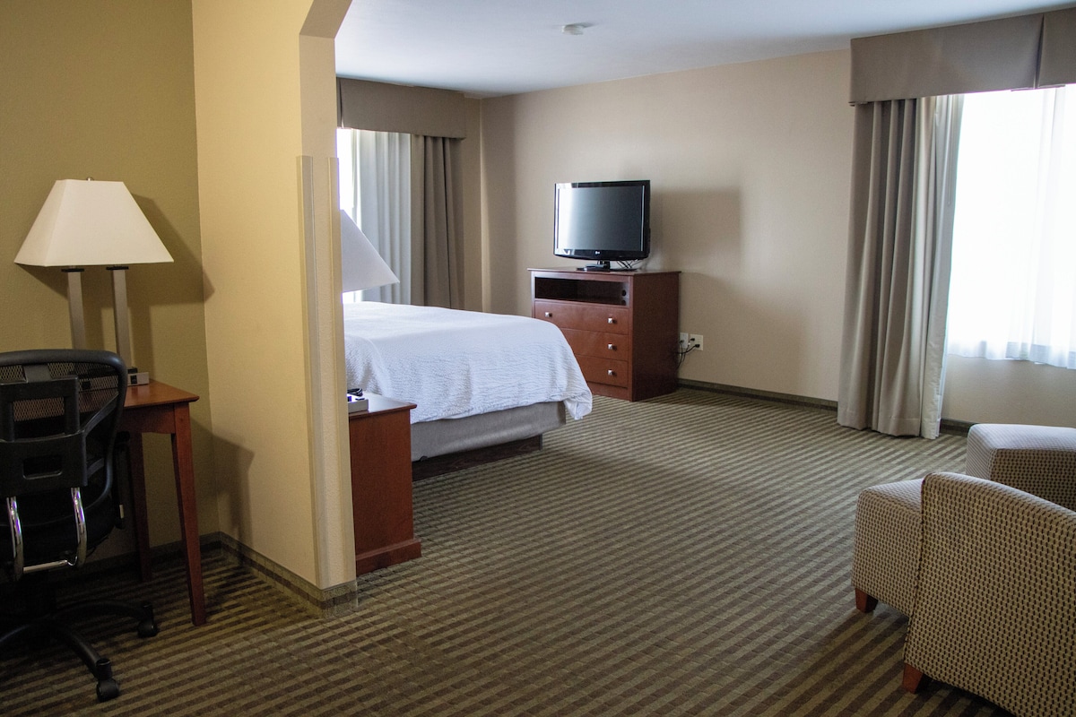Quarto de hotel com cama, televisão sobre um móvel de madeira, escrivaninha com abajur e uma cadeira.