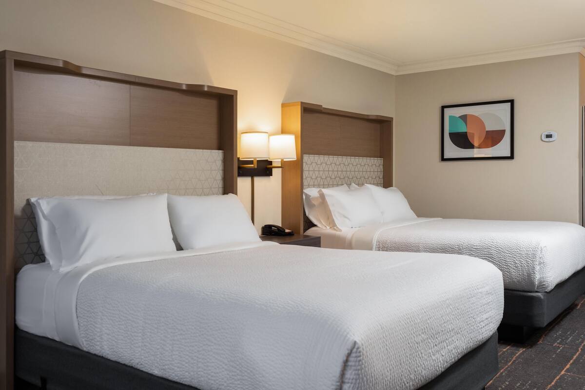 Quarto de hotel com duas camas, uma mesa de cabeceira, uma luminária e uma obra de arte na parede.