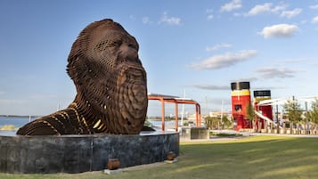 Uma grande escultura metálica da cabeça de uma baleia em um parque público, com céu claro e edifícios ao longe.