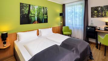 Quarto de hotel com uma cama grande, duas cadeiras, uma mesa com computador e duas luminárias de parede.