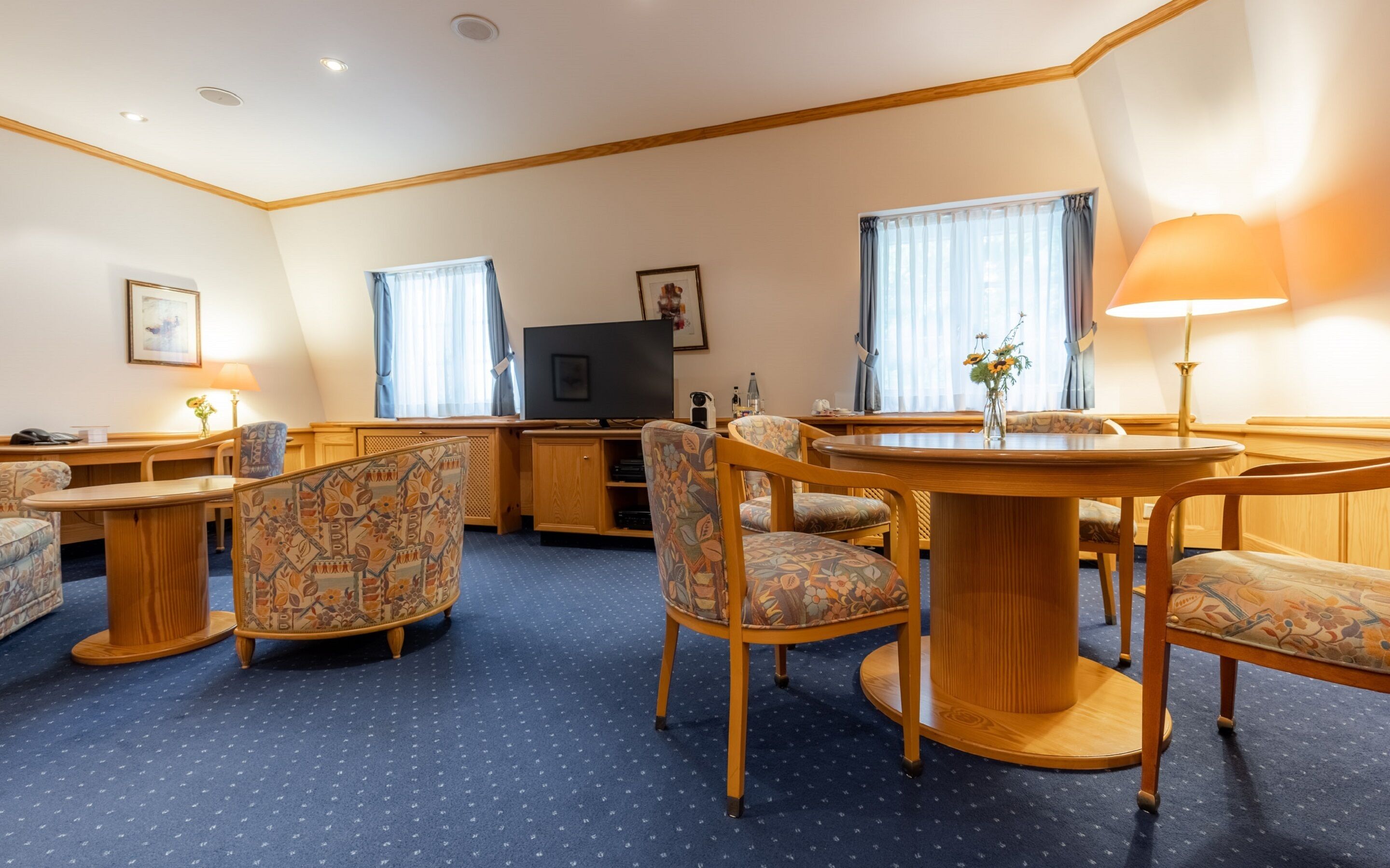 Quarto de hotel com área de refeições, destacando uma mesa redonda de madeira e cadeiras com padrão floral. Há uma televisão sobre um armário de madeira, um sofá e uma janela com cortinas.
