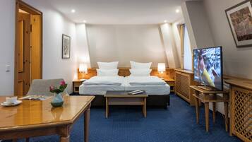 Quarto de hotel com cama de casal, mesas de cabeceira com luminárias, televisão, escrivaninha de madeira, cadeira e um vaso com flores.