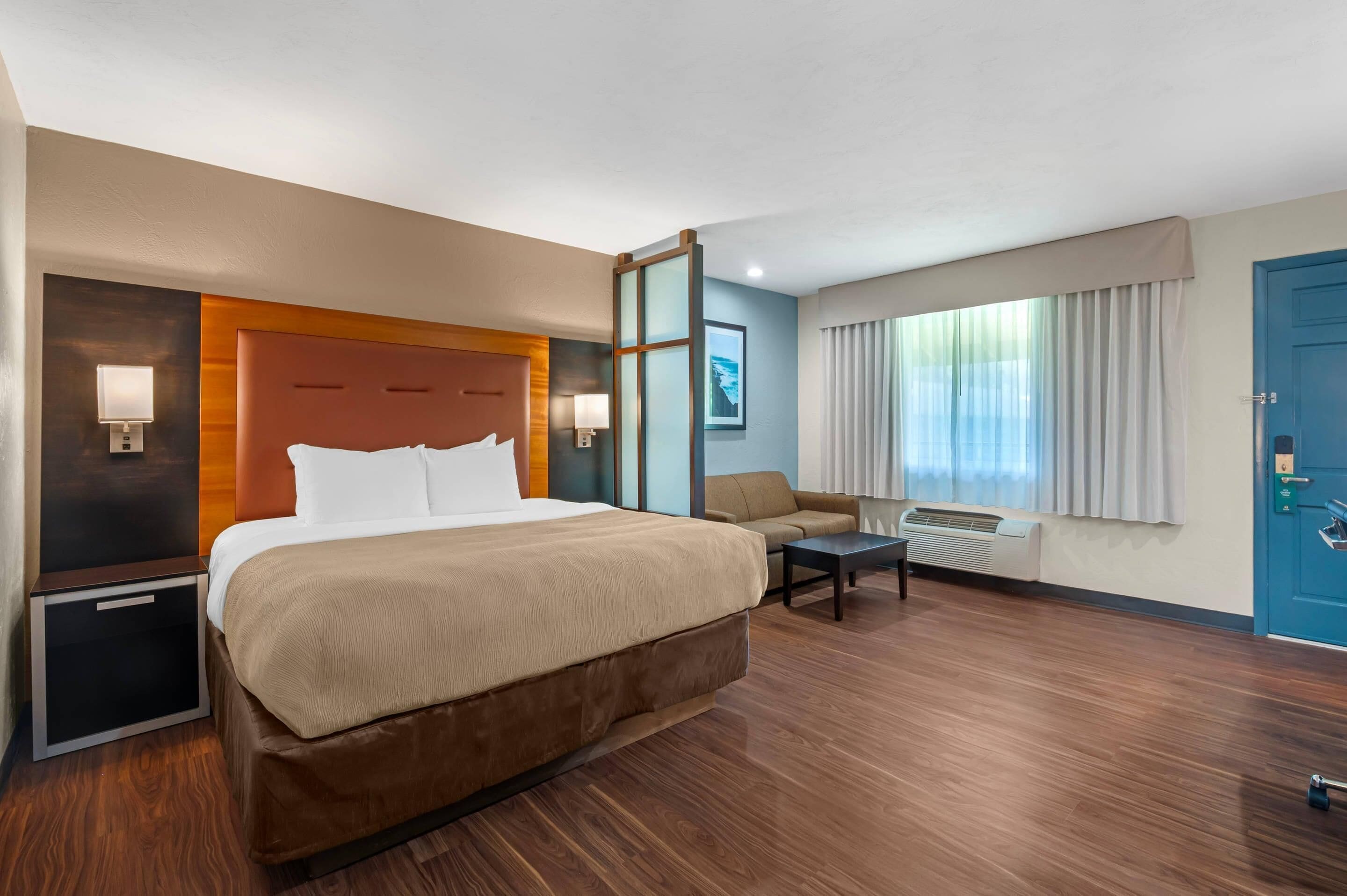 Quarto de hotel com uma cama grande, um sofá, uma mesa pequena, uma mesinha de cabeceira, uma janela com cortinas e uma porta.