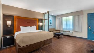 Quarto de hotel com uma cama grande, um sofá, uma mesa pequena, uma mesinha de cabeceira, uma janela com cortinas e uma porta.